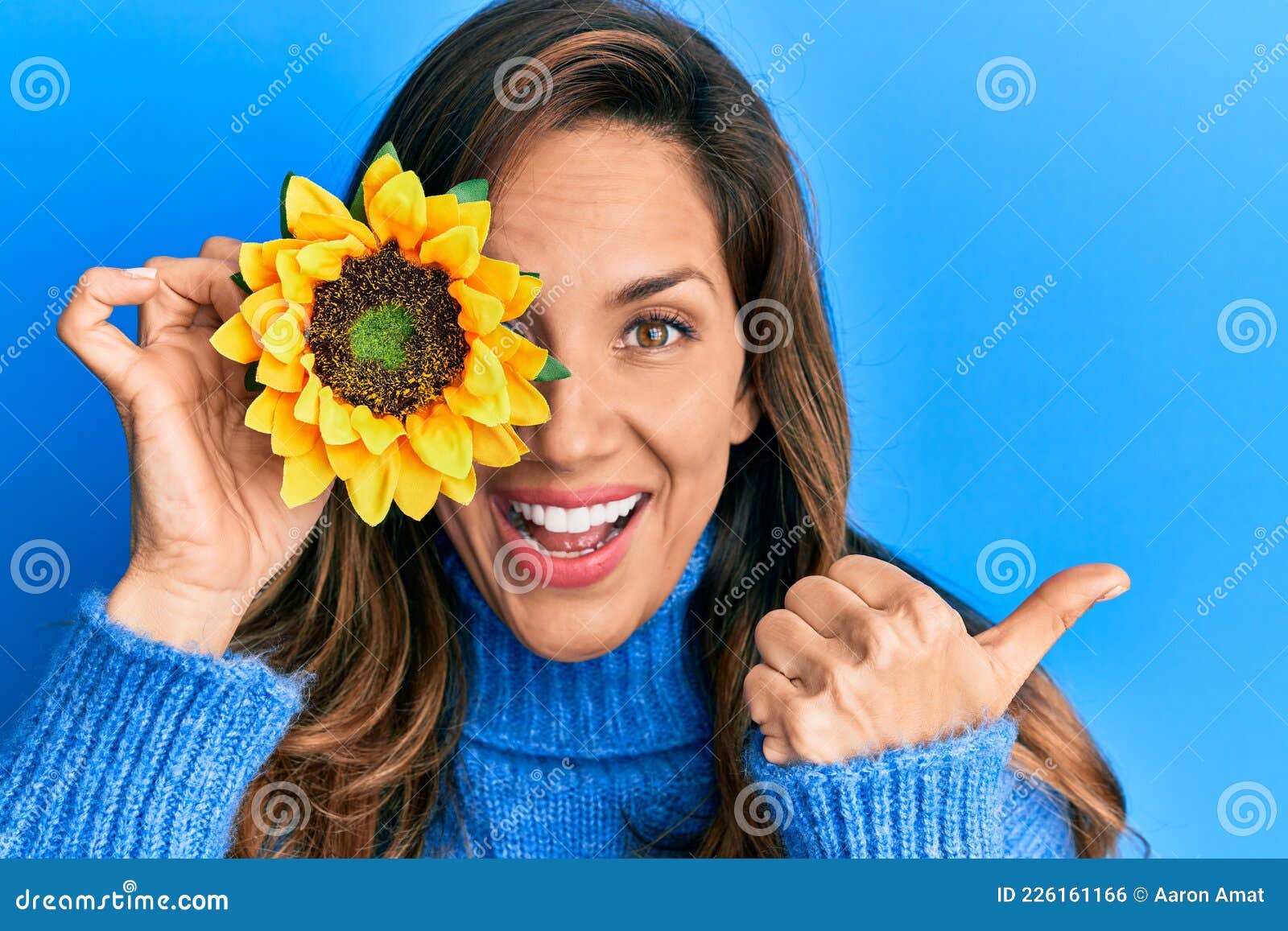 Young Latin Woman Holding Sunflower Over Eye Pointing Thumb Up To The
