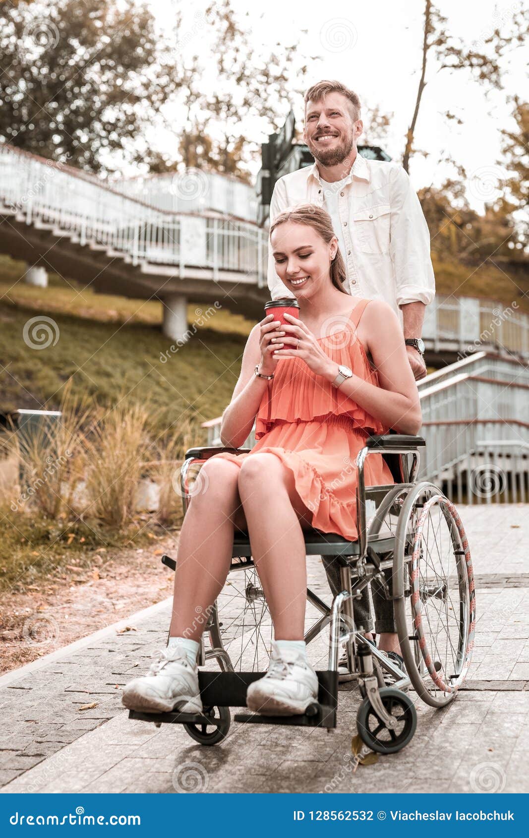 Young Man Pushing the Wheelchair and His Girlfriend Holding Glass of ... Young Man Pushing the Wheelchair and His Girlfriend Holding Glass of ...