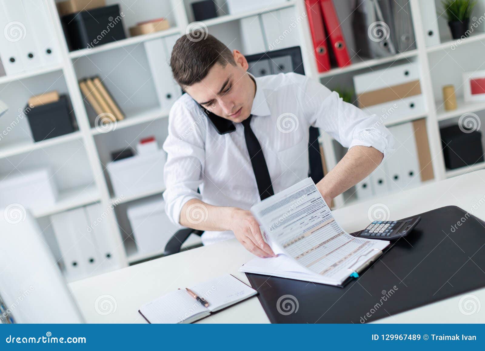 A Young Man Sitting at a Table in the Office, Talking on the Phone and ...