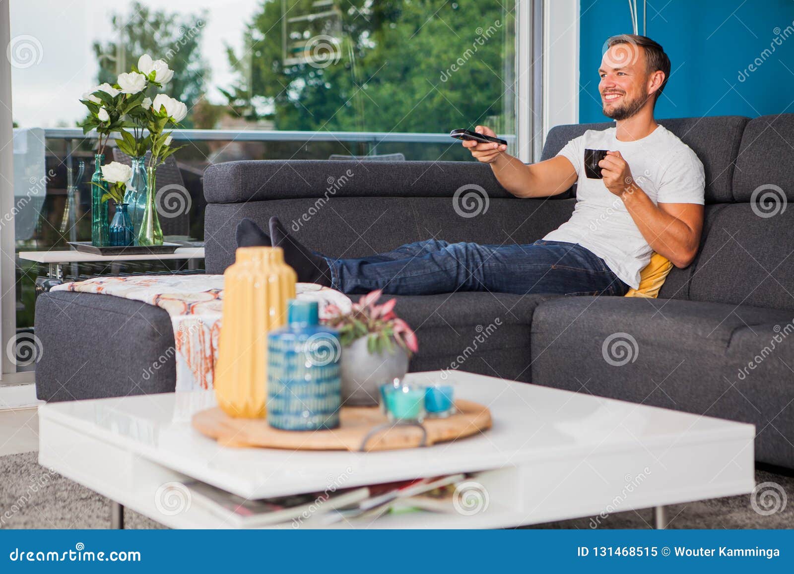 Young Man Using a Remote Control while Drinking Coffee on the Couch ...