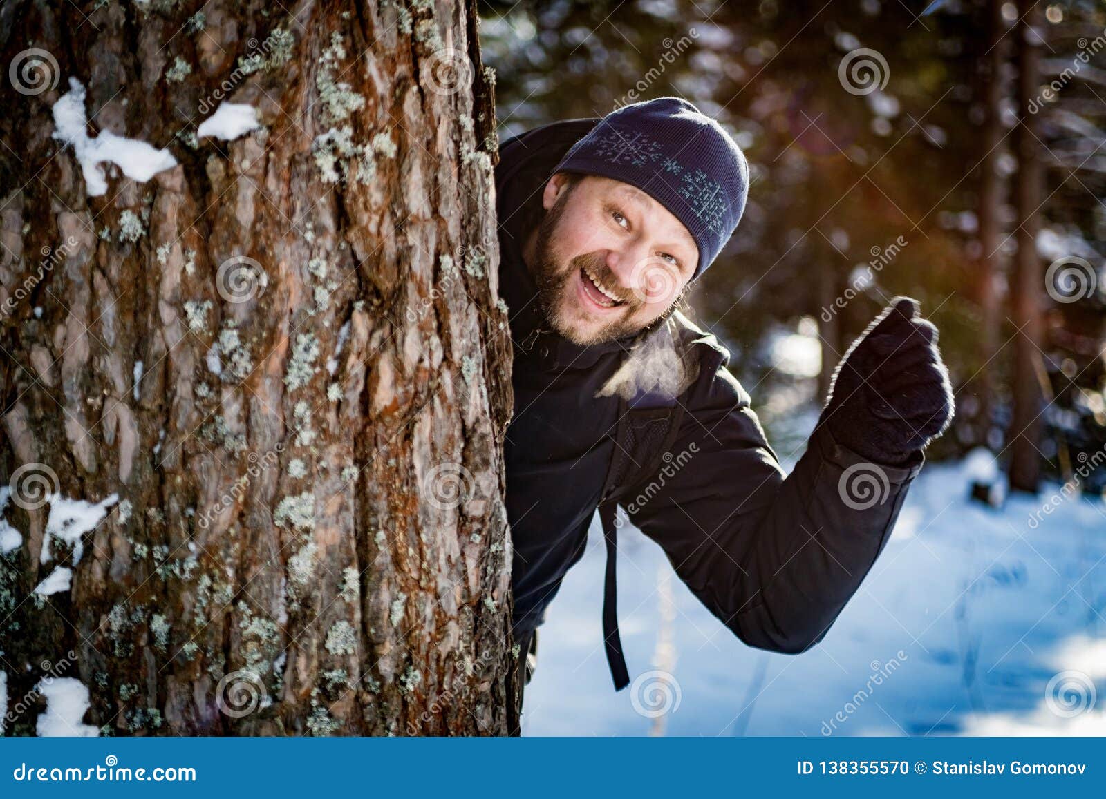 A Young Man in a Winter Forest Looks Out from Behind a Tree and Waves ...