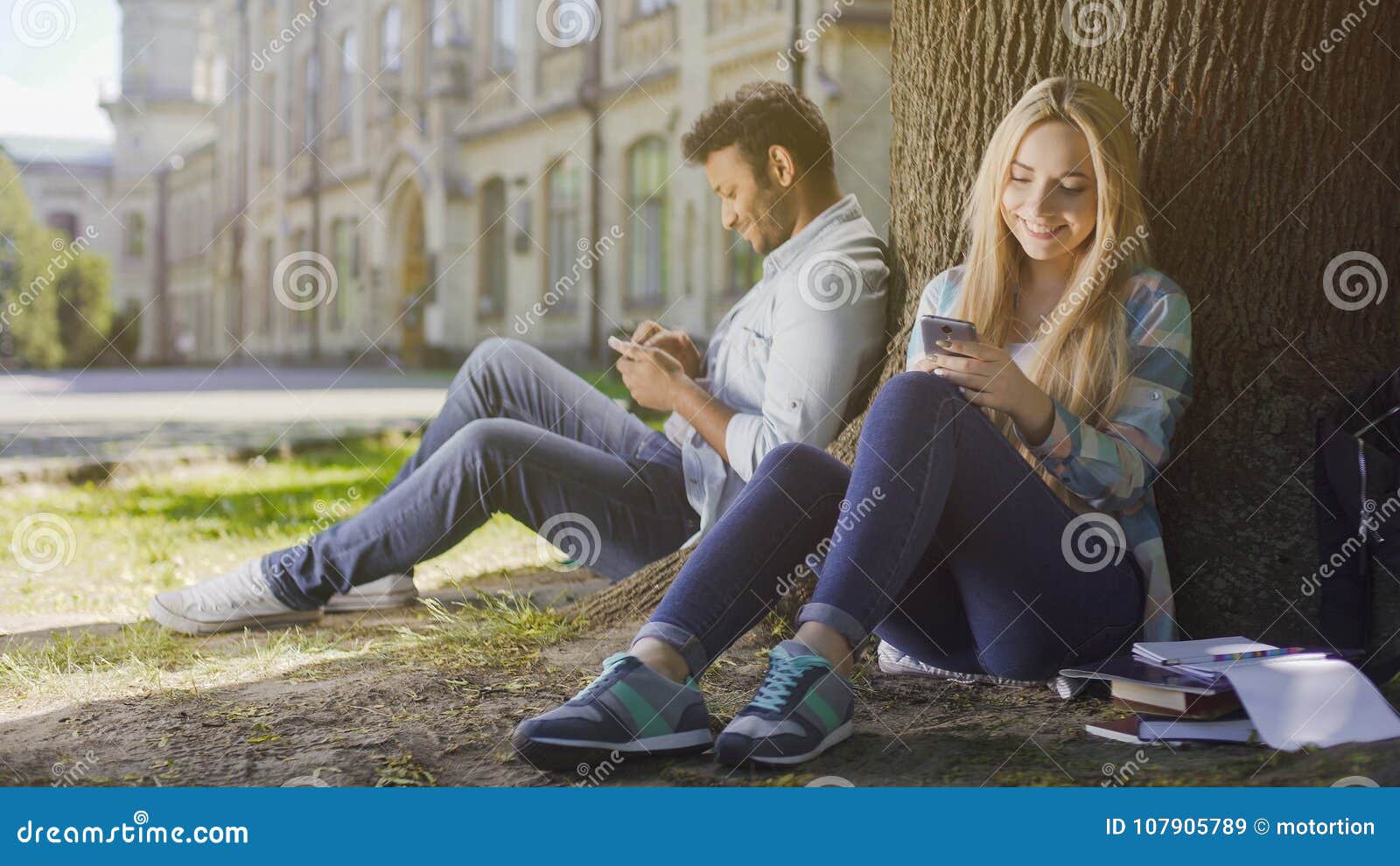 Young People Sitting Under Tree, Using Cellphone, Smiling To Themselves ...