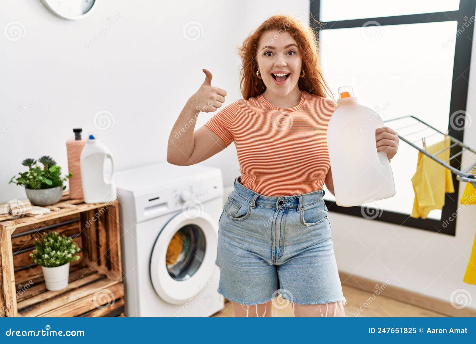 Young Redhead Woman Doing Laundry Holding Detergent Bottle Smiling Happy And Positive Thumb Up