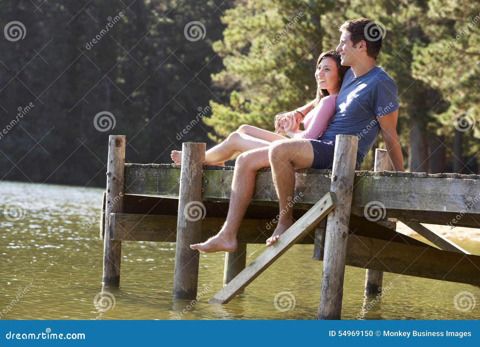 Young Romantic Couple Sitting On Wooden Jetty Looking Out Over Lake