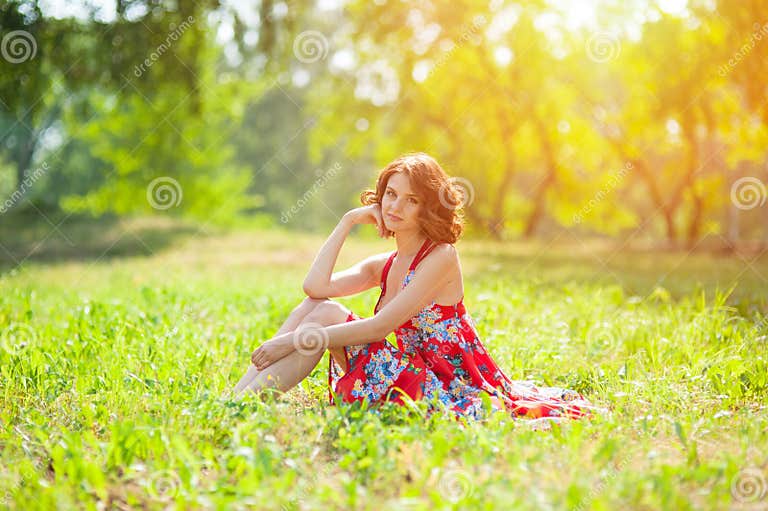 Young Brunette Girl In Red Dress Posing On Grass In Summer Park In The