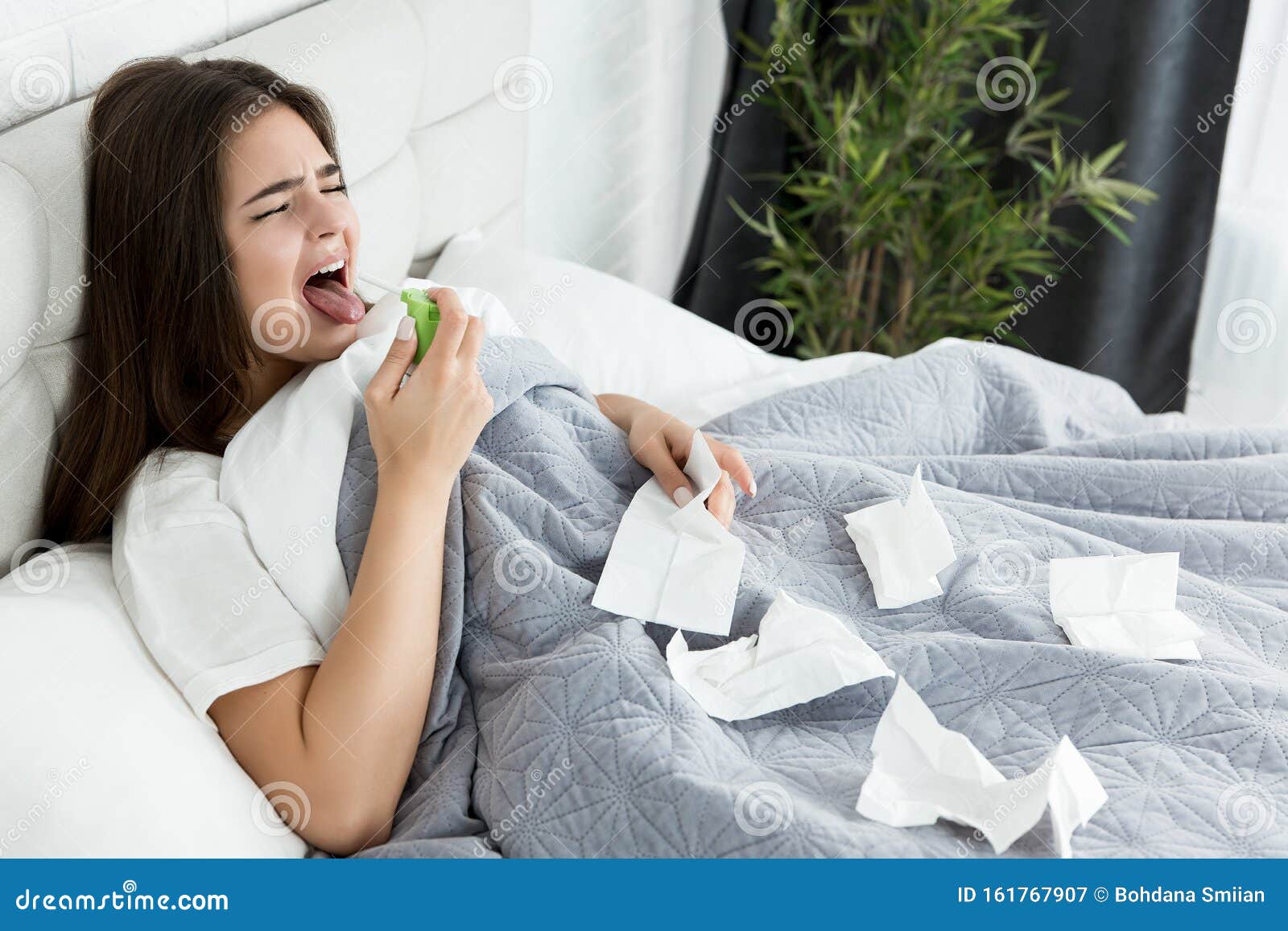 Young Sick Woman Sitting In Bed Spraying Medicine Into Her Sore Throat