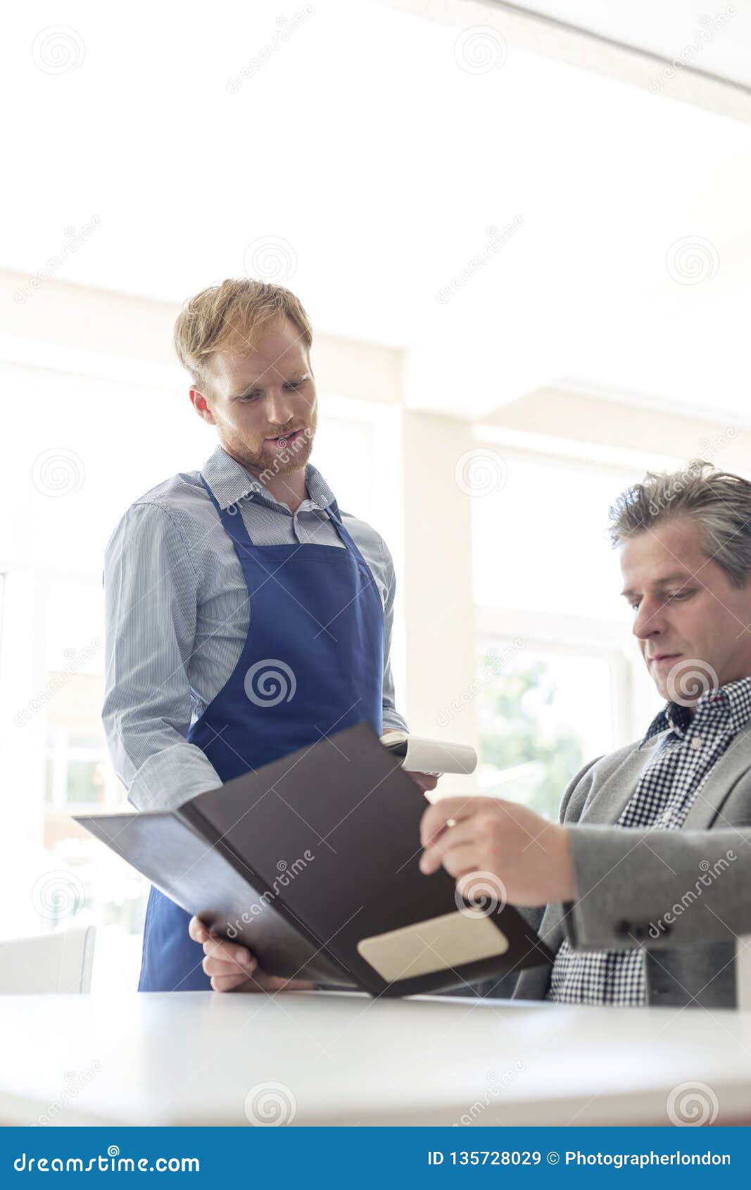 Young Waiter Standing by Customer Reading Menu at Table in Restaurant ...