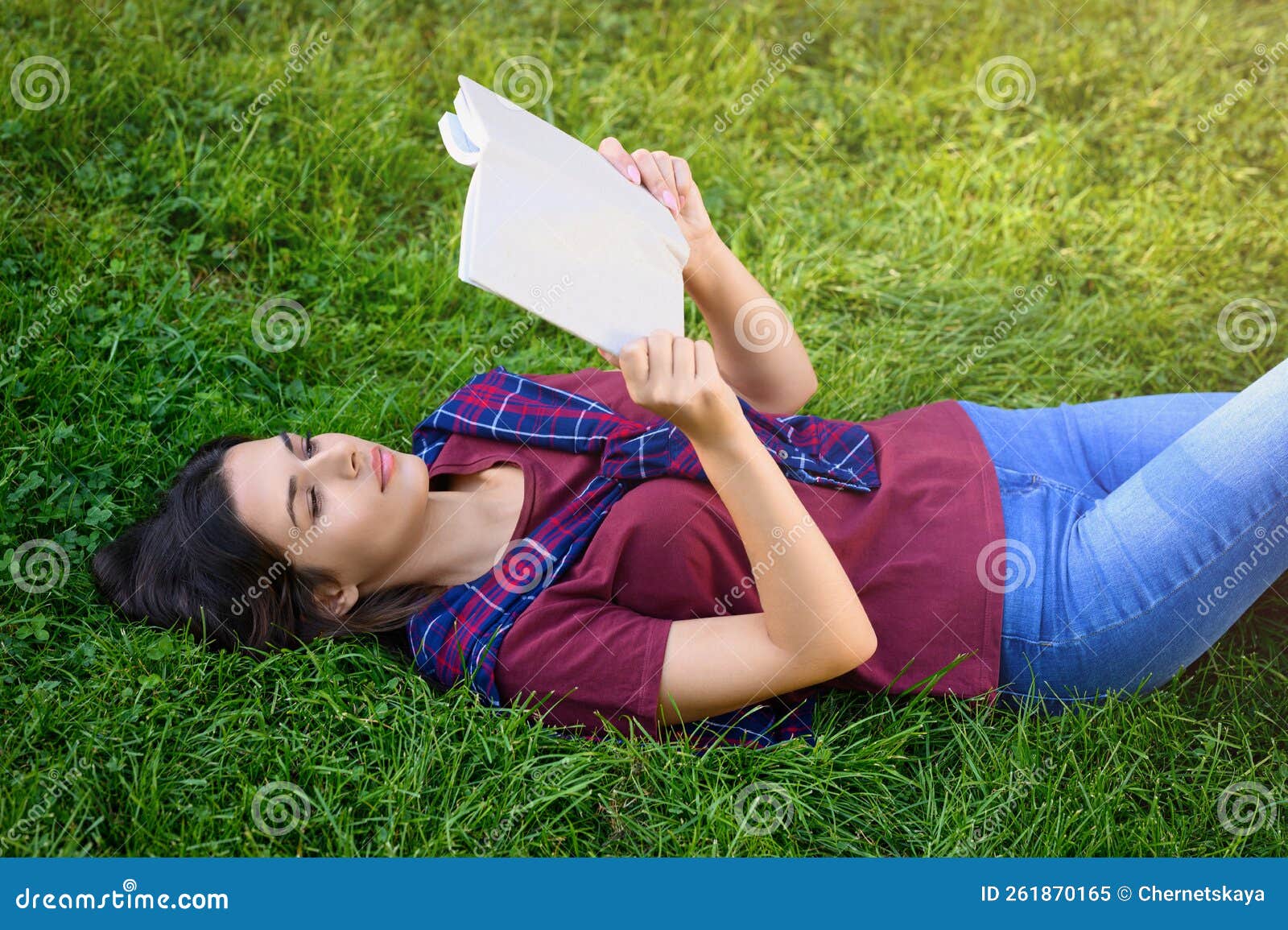 Young Woman Reading Book While Lying On Green Grass Outdoors Stock