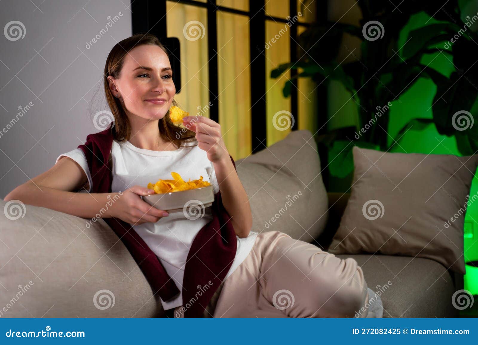Young Woman is Relaxing on the Couch, Watching TV and Eating Chips ...