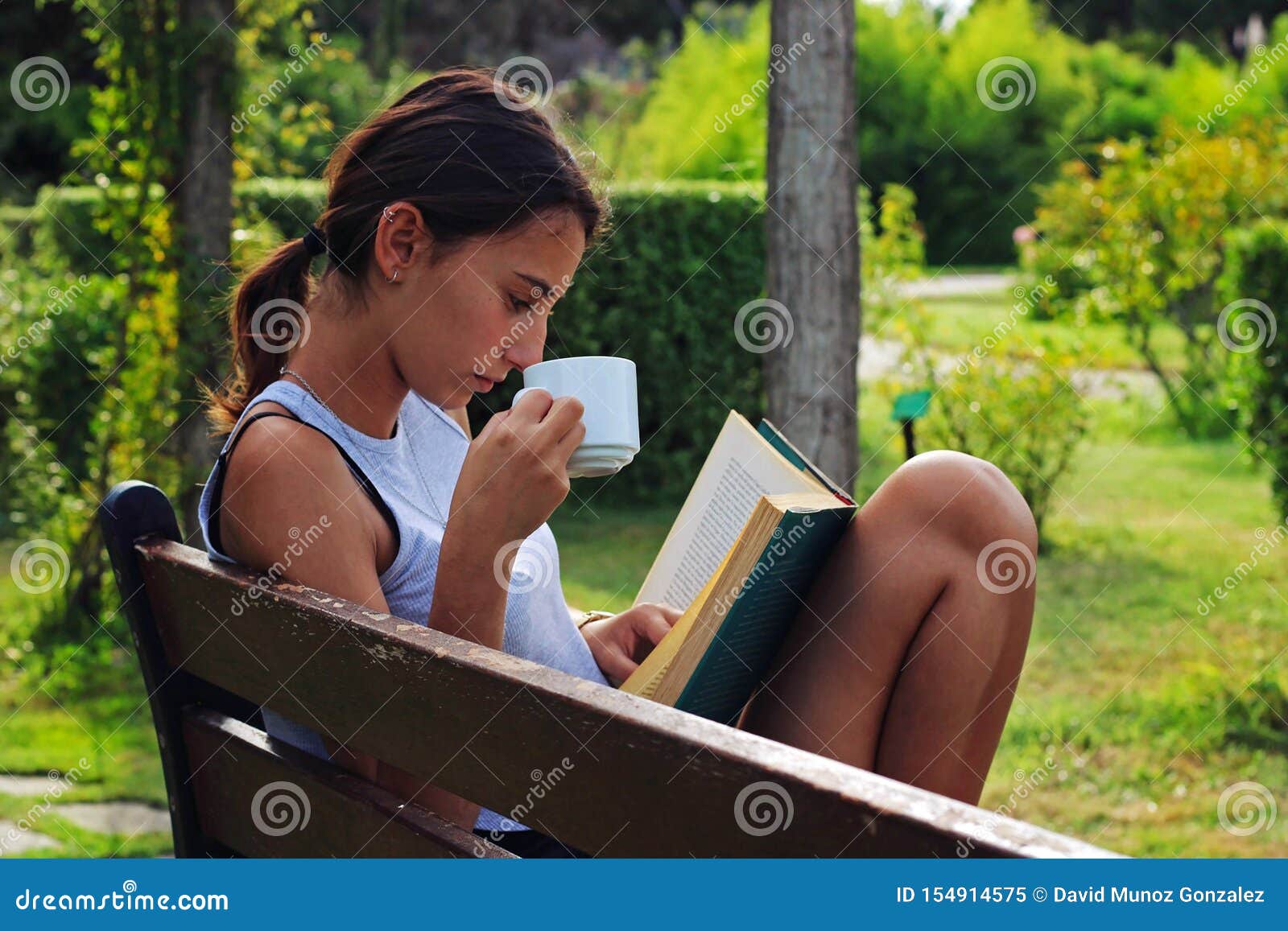Woman Sitting On A Bench Reading A Book In The Woods And Drinking A Cup