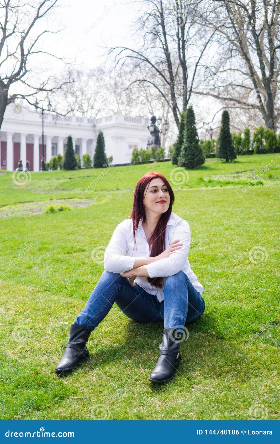 Young Woman In White Shirt And Blue Jeans Sitting On A Green Grass