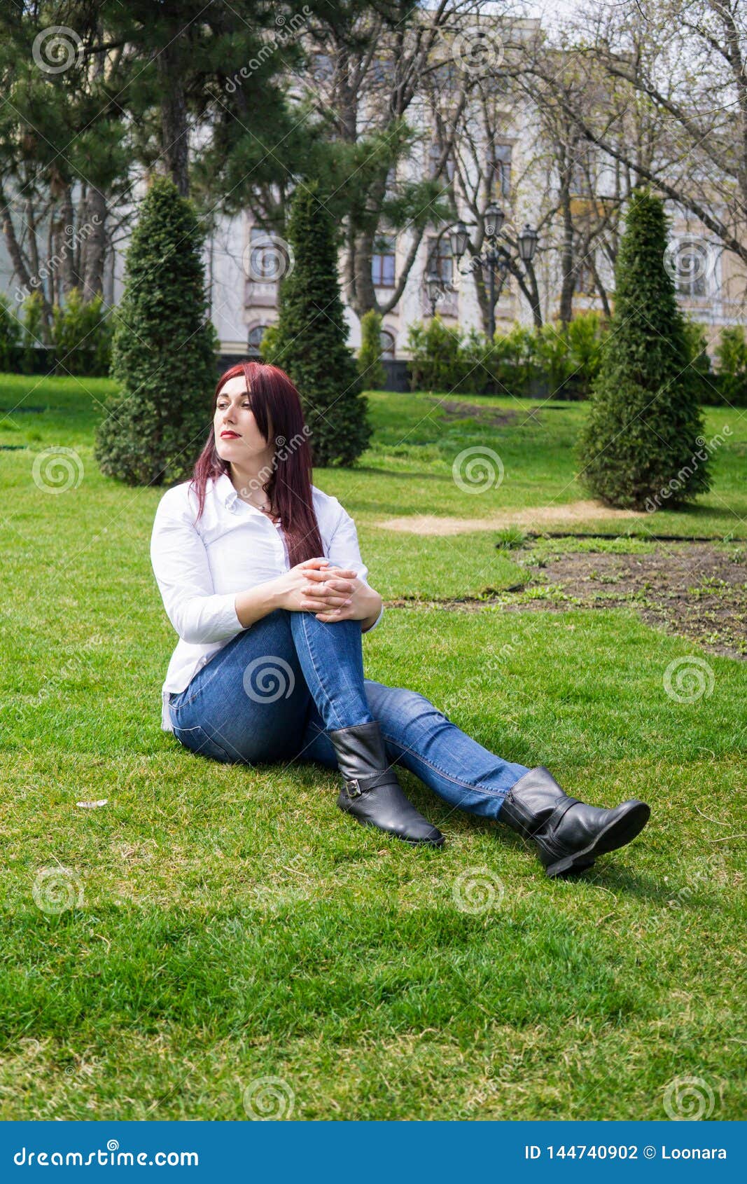 Young Woman In White Shirt And Blue Jeans Sitting On A Green Grass