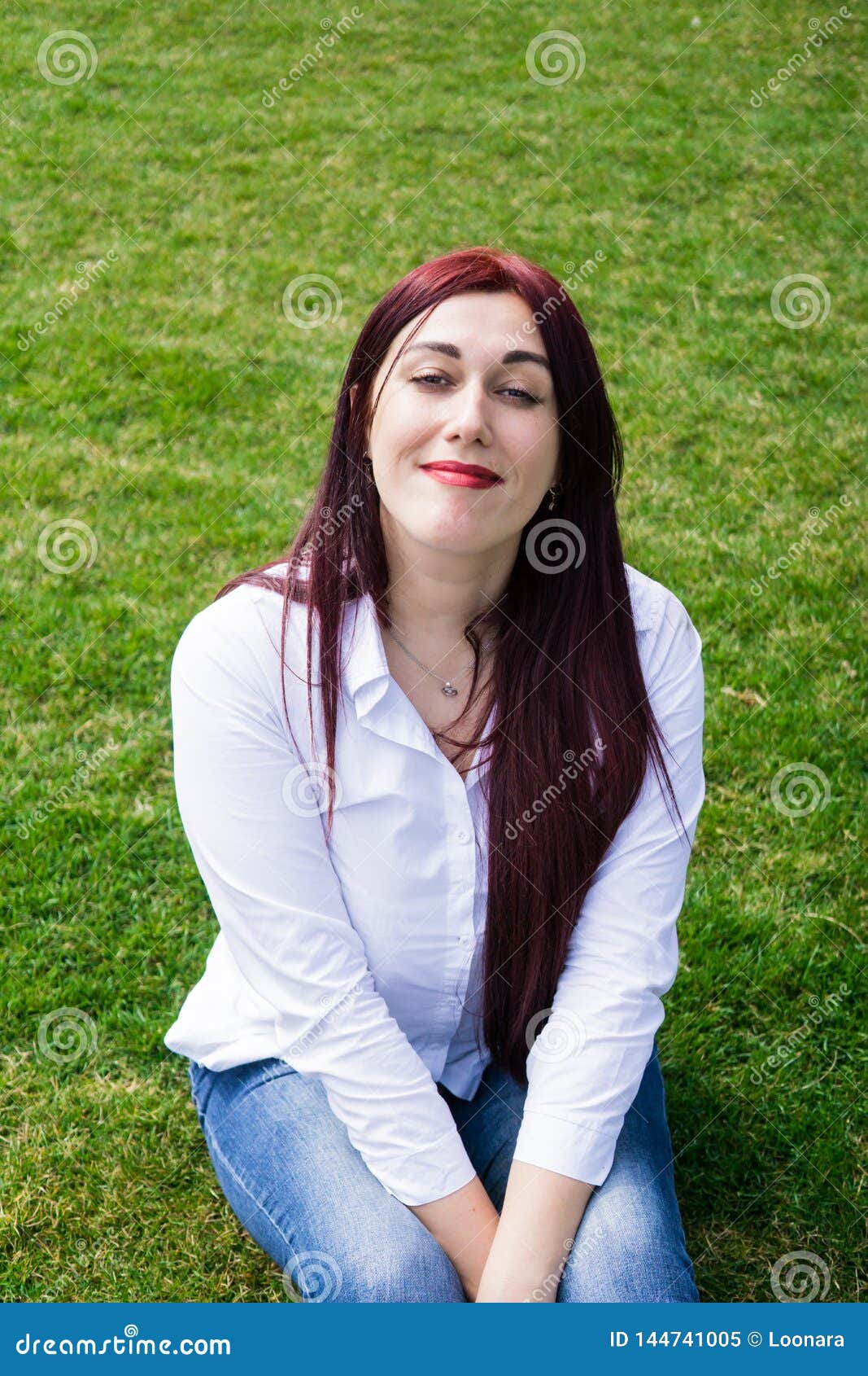 Young Woman In White Shirt And Blue Jeans Sitting On A Green Grass