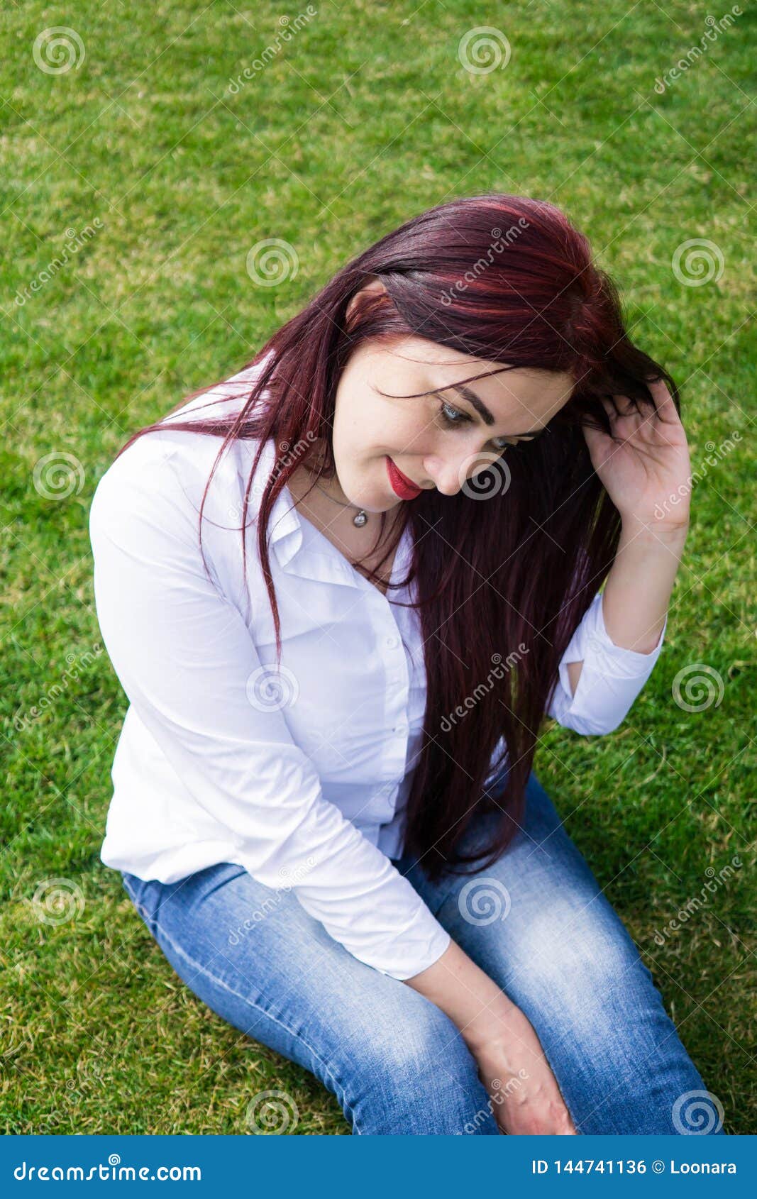 Young Woman In White Shirt And Blue Jeans Sitting On A Green Grass