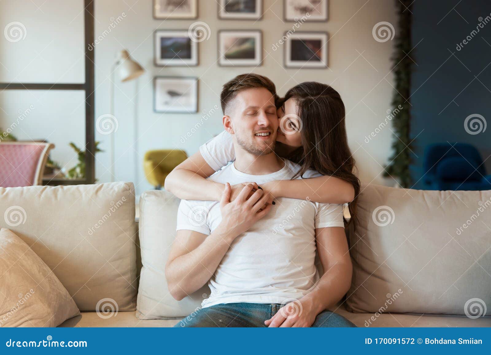Young Woman Embracing Her Husband`s Shoulders And Kisses Him While He