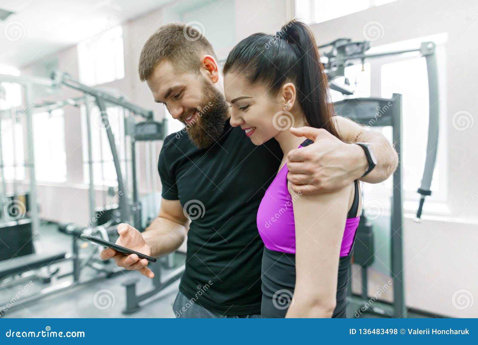 Young Woman Talking With Personal Trainer In Gym Looking In Smartphone